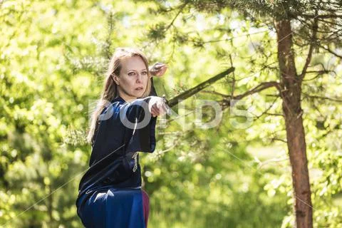 A female warrior with a sword poses on a stump in the forest. Wushu ...