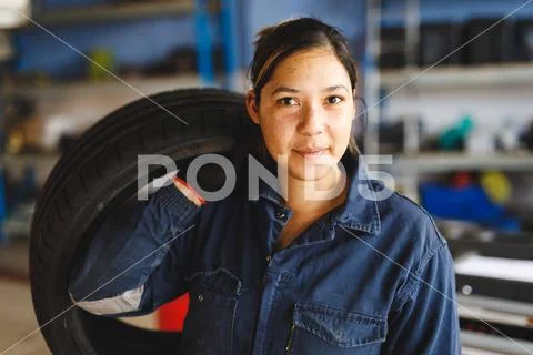 Mixed race female car mechanic wearing overalls, holding tire, looking ...