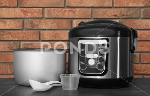 Modern electric multi cooker, parts and accessories on table near brick ...