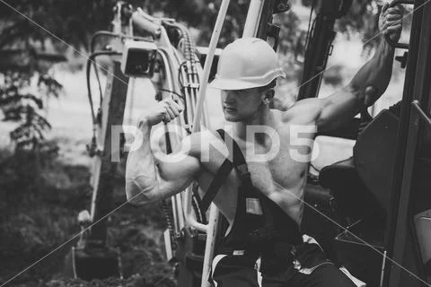 Muscular builder in hard hat shows biceps, poses. ~ Hi Res #104371083