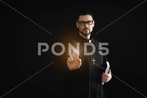 Priest with Bible making blessing gesture and holy light on dark ...